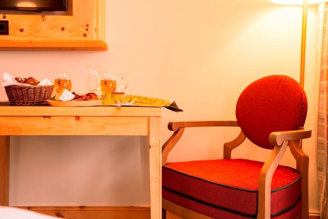 Uma mesa de madeira com uma cesta de petiscos, duas chávenas de chá e um prato de comida. Uma cadeira estofada vermelha está posicionada ao lado da mesa.
