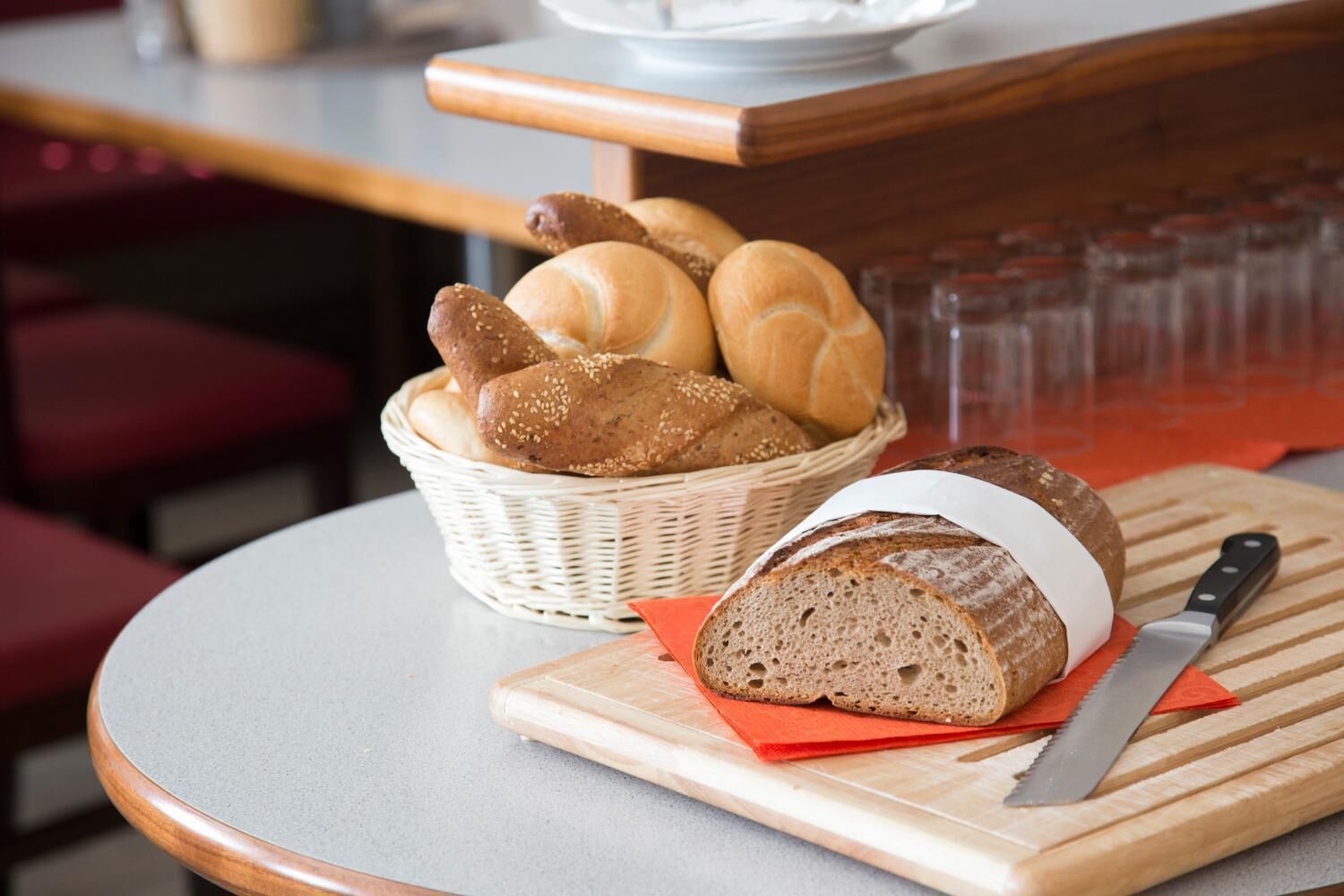 Uma mesa redonda com uma cesta de pães variados e uma fatia de pão em uma tábua de corte.