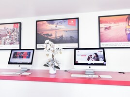 Bancada de recepção com três monitores exibindo conteúdo promocional da Red Planet Hotels, um vaso com flores brancas e superfície da mesa vermelha.
