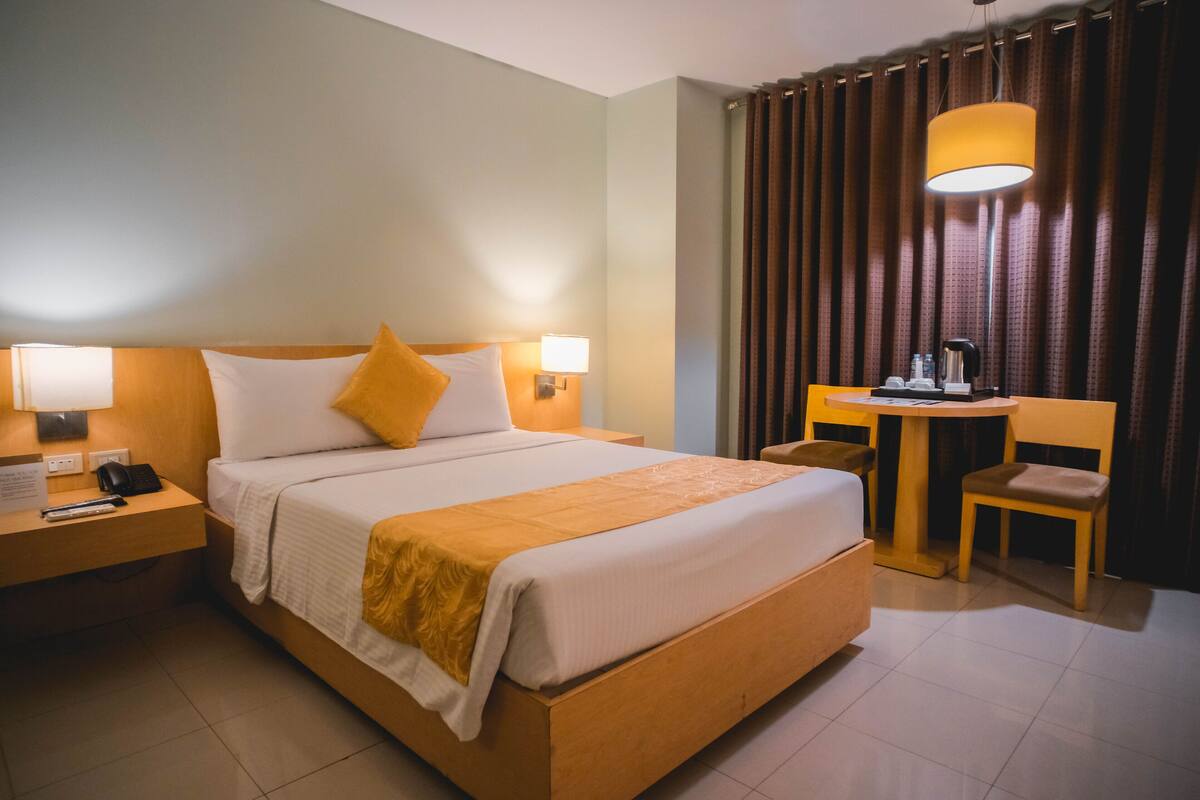 Quarto de hotel com uma cama grande, mesas de cabeceira, uma escrivaninha com cadeira e uma luminária.