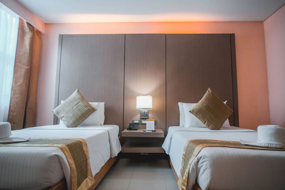Quarto de hotel com duas camas, mesa de cabeceira com abajur e cabeceira de madeira.