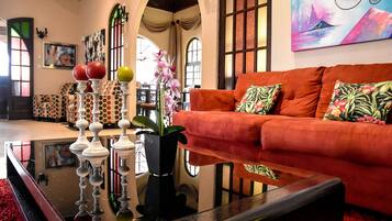 Sala de estar com um sofá vermelho, uma mesa de centro preta e um vaso com flores. Há janelas com cortinas, um quadro na parede e um sofá estampado ao fundo.