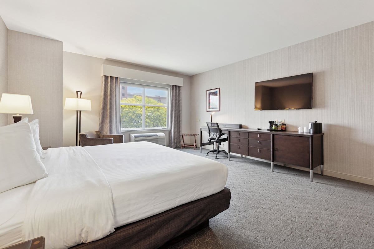 Quarto de hotel com uma cama grande, uma escrivaninha com cadeira, TV de tela plana na parede e uma janela com cortinas.