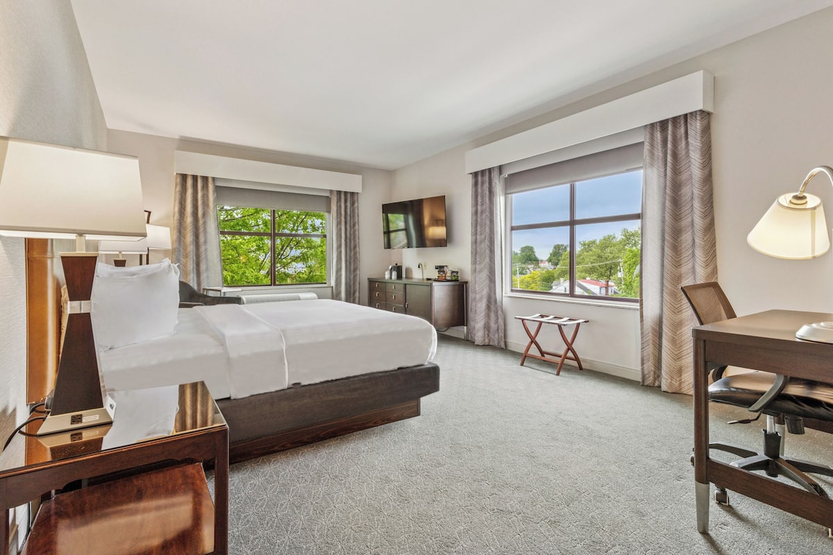 Quarto de hotel com cama, escrivaninha, cadeira, televisão e amplas janelas com cortinas.