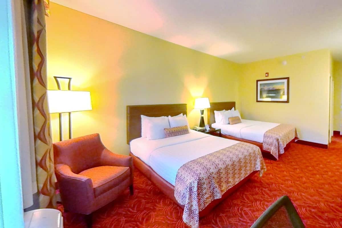 Quarto de hotel com duas camas, uma cadeira, um abajur, um quadro na parede e piso coberto por carpete.