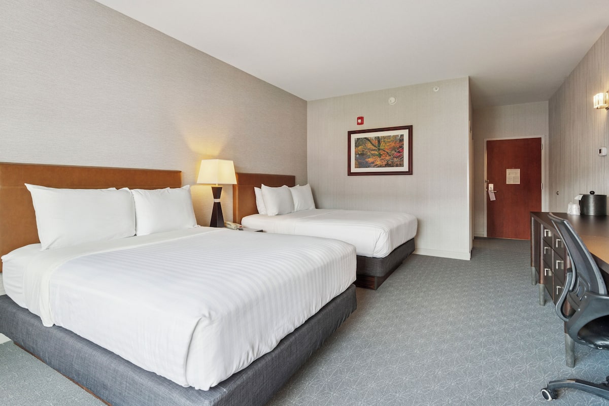 Quarto de hotel com duas camas, uma escrivaninha, uma cadeira, um abajur, um quadro na parede e uma porta.