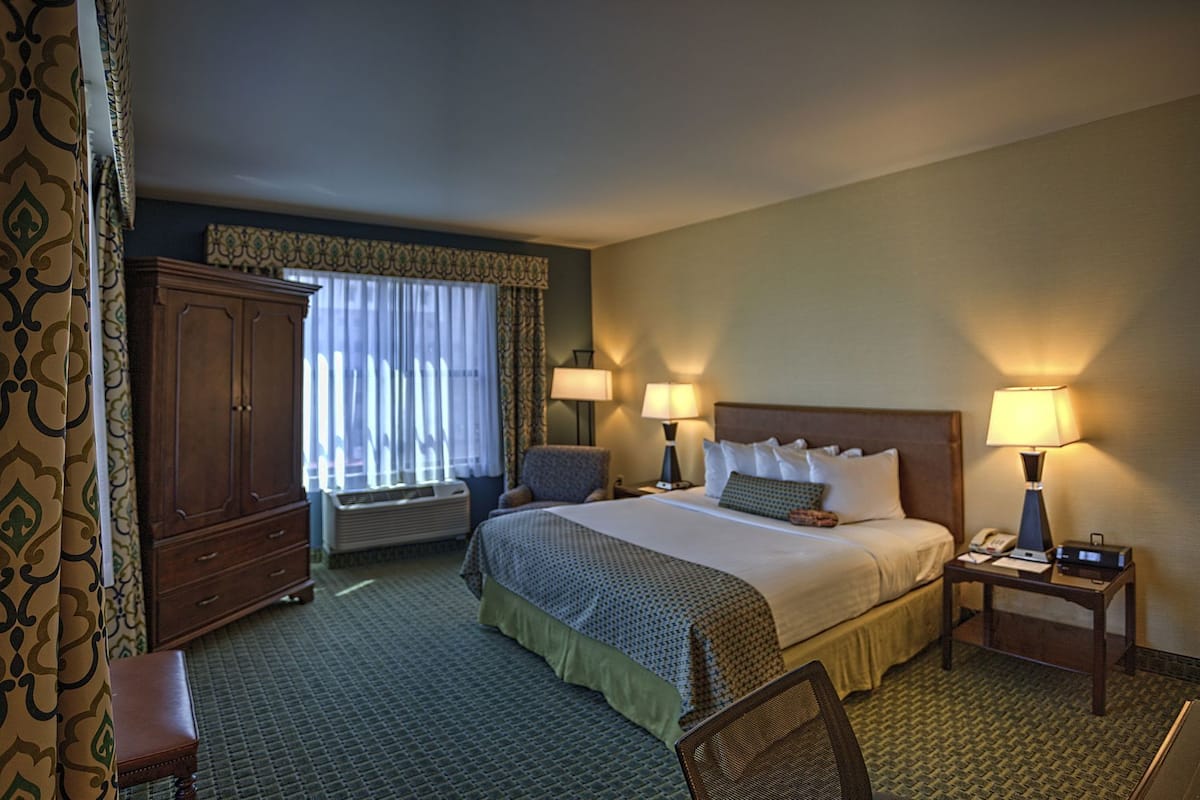 Quarto de hotel com uma cama, duas mesas de cabeceira com luminárias, uma cadeira, um guarda-roupa e uma janela com cortinas.