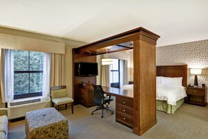 Quarto de hotel com uma cama grande, uma mesa com cadeira, televisão, luminária e janela com cortinas.