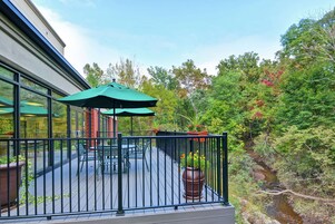 Terraço com móveis de exterior e guarda-sóis, rodeado por uma grade de metal e vista para uma área arborizada.