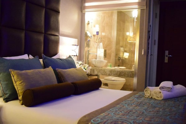 Quarto de hotel com uma cama grande, banheiro com box de vidro e cama bem arrumada com toalhas.