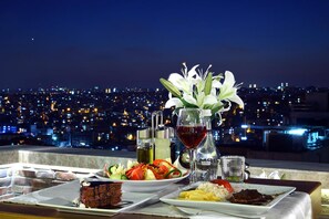 Uma mesa posta com sobremesa, salada e pratos de comida, com vista para o horizonte urbano ao fundo.