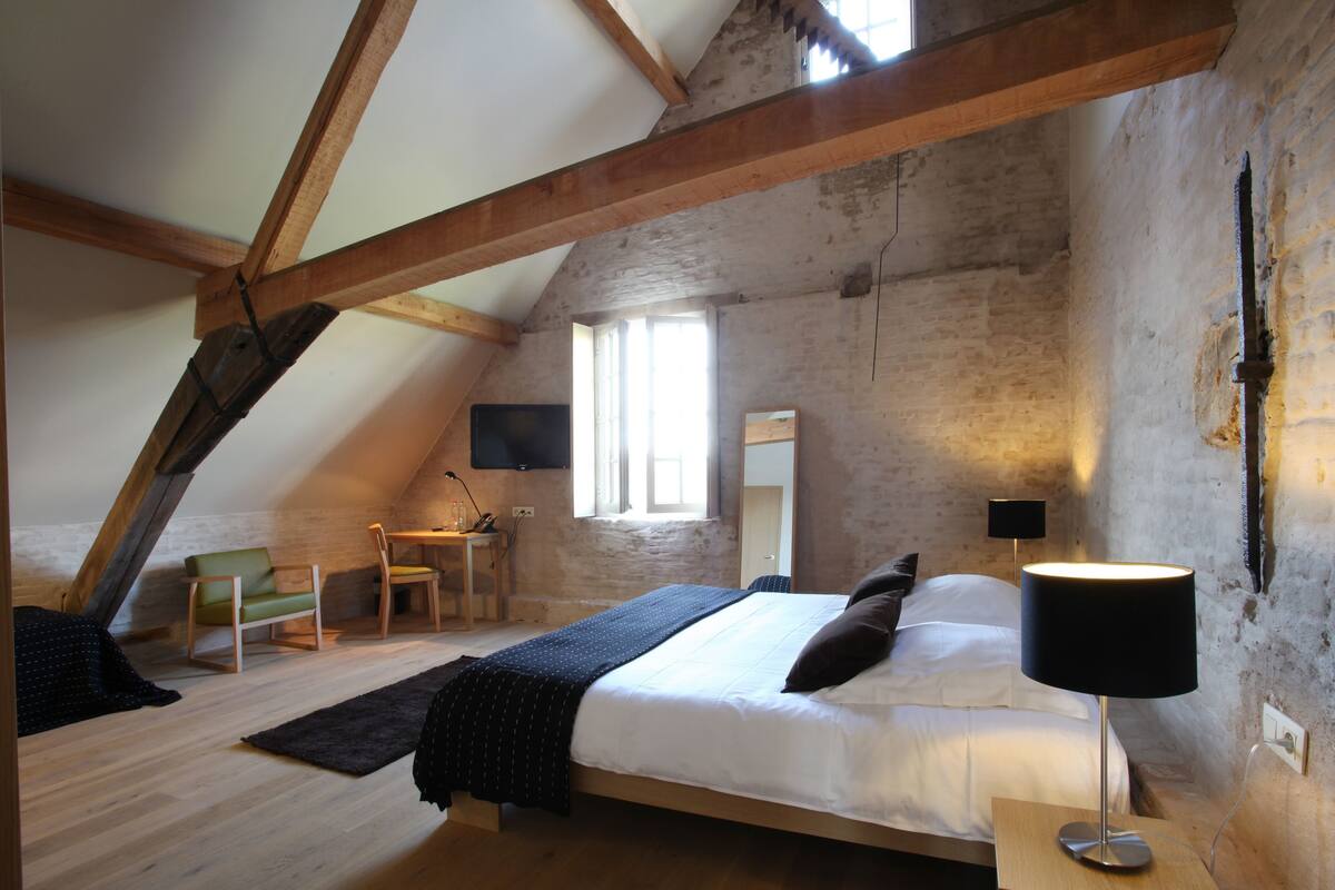 Quarto com teto inclinado, vigas de madeira, cama com roupa de cama branca, escrivaninha com cadeira, televisão e janela com luz natural.