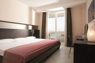 Quarto de hotel com uma cama, uma escrivaninha com luminária, uma cadeira e vista para o exterior através de uma grande janela.
