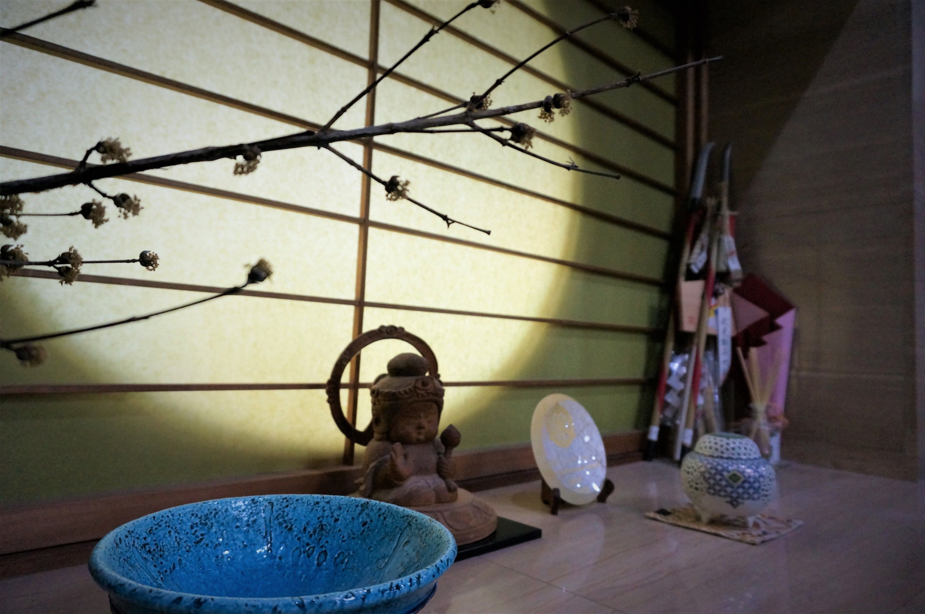 Um quarto com piso de esteira de tatame, uma tigela de cerâmica azul, uma estátua de pedra e um objeto decorativo branco sobre um suporte.