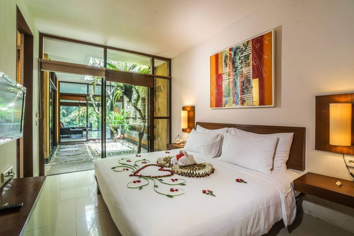 Quarto de hotel com cama, televisão, escrivaninha e vista para um jardim através de portas de vidro.