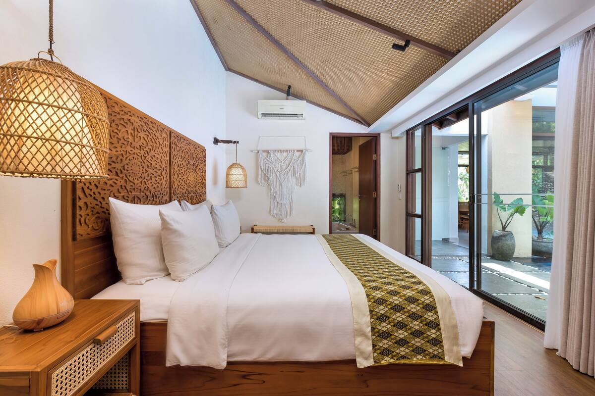 Um quarto espaçoso com uma cama grande, uma mesinha de cabeceira de madeira, uma luminária de fibra natural, um painel de macramê na parede e vista para o exterior através de amplas janelas.
