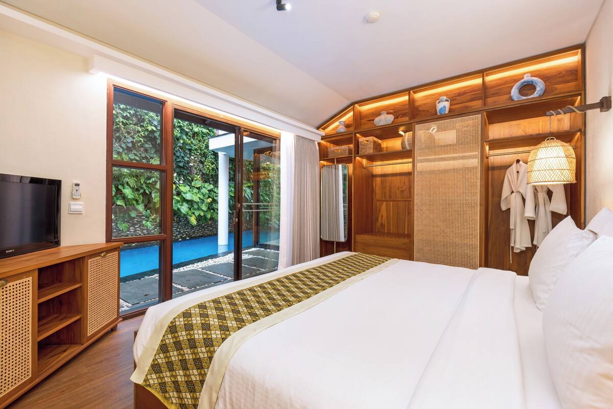 Quarto de hotel com uma cama grande, televisão, armário de madeira e porta de vidro deslizante para o exterior.