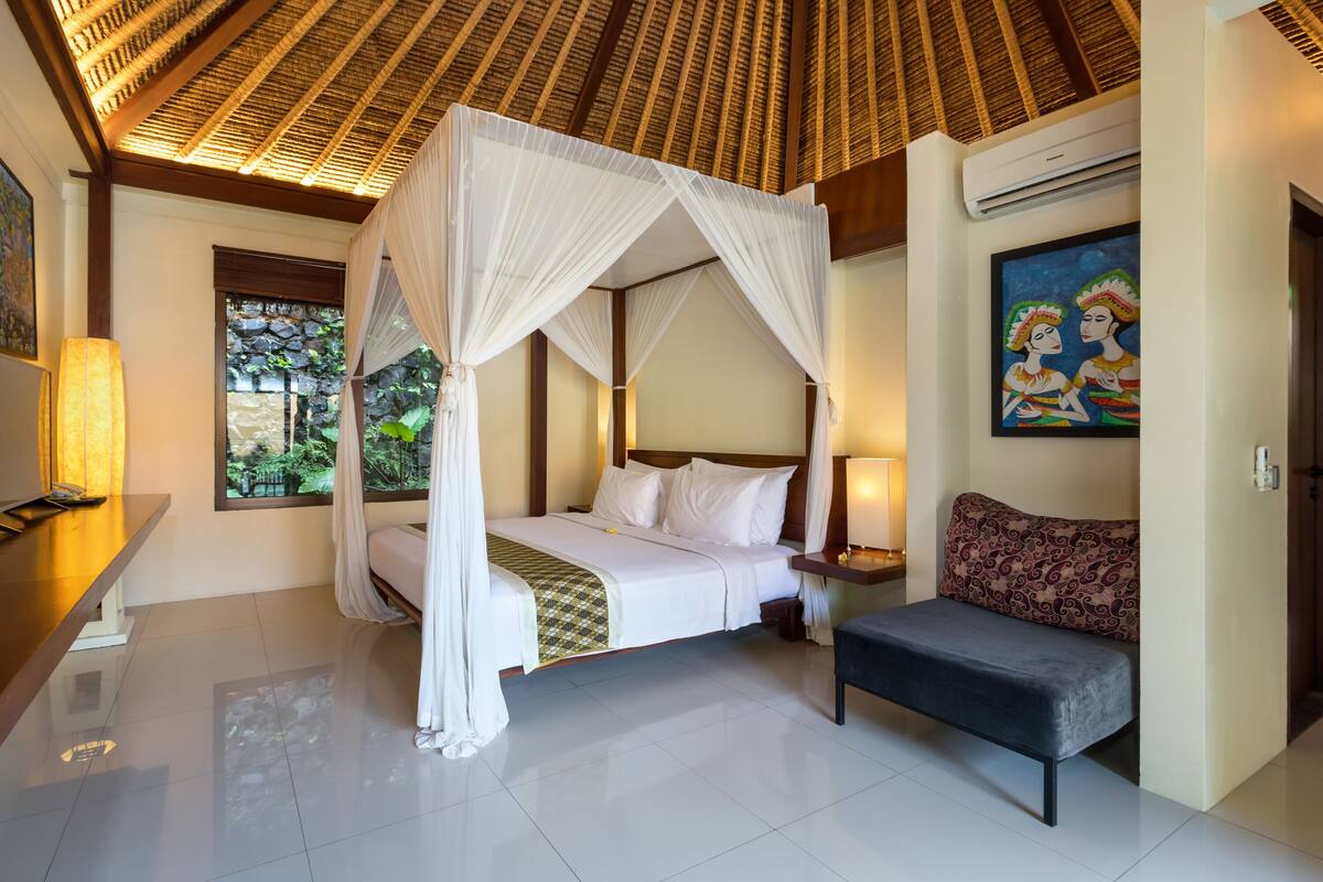 Quarto com cama de dossel, um banco, um quadro e janela com vista para vegetação.
