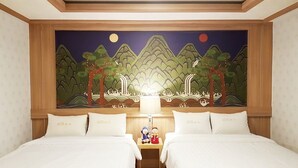 Quarto de hotel com duas camas, uma mesa de cabeceira com abajur e um grande painel na parede retratando uma paisagem montanhosa.