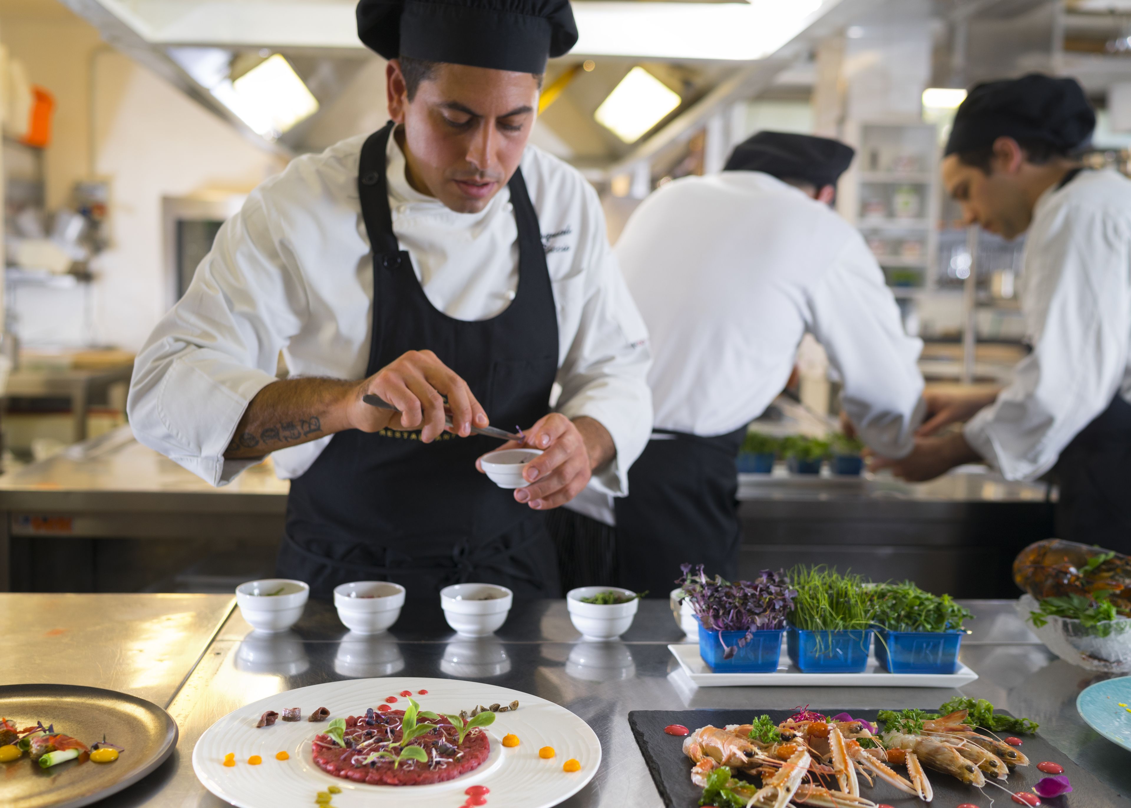Um chef preparando alimentos em uma cozinha profissional, com diversos pratos e ingredientes sobre o balcão.
