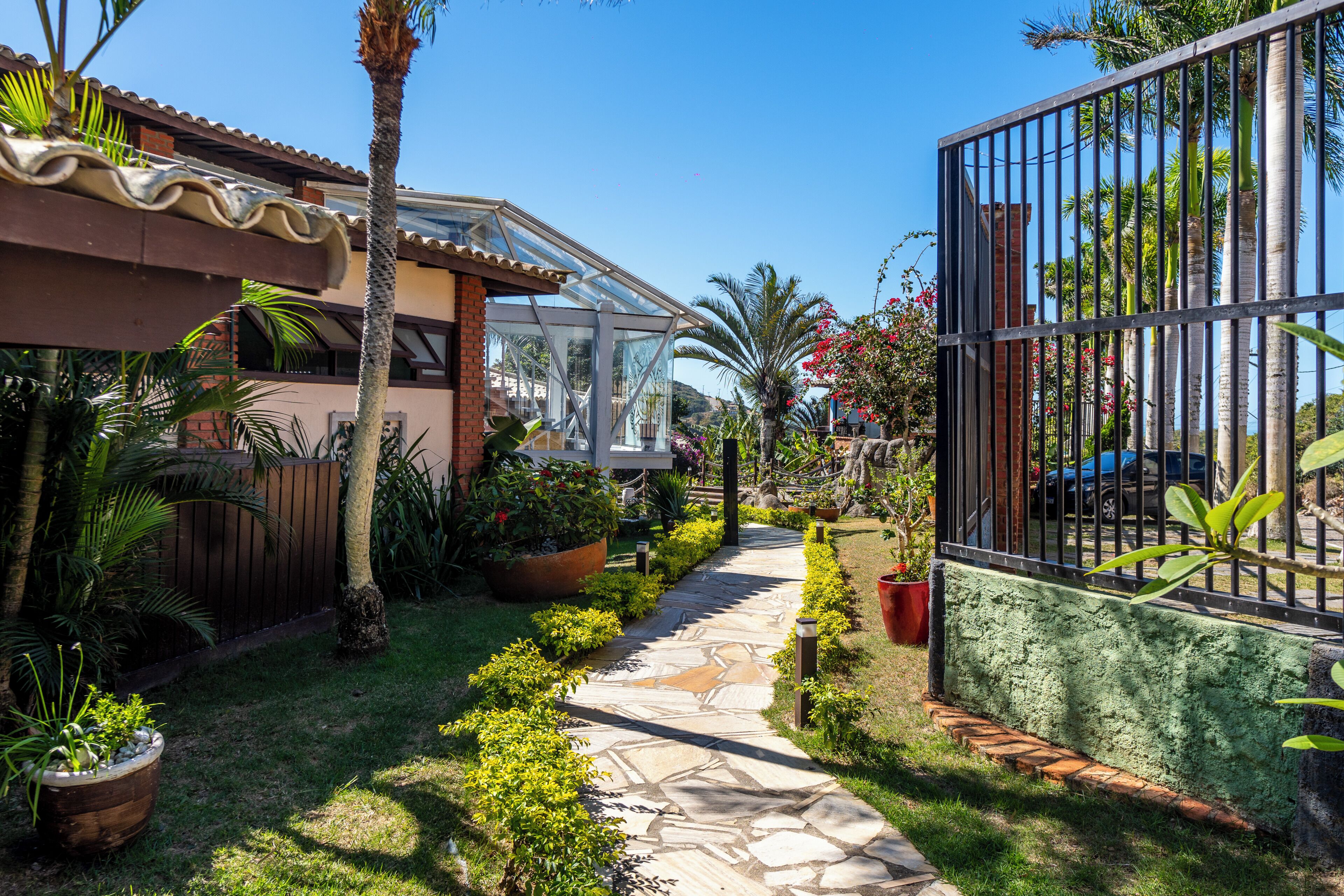 Uma passagem no jardim leva a uma casa com conservatório de vidro, cercada por plantas em vasos e um portão de metal.