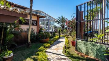 Uma passagem no jardim leva a uma casa com conservatório de vidro, cercada por plantas em vasos e um portão de metal.