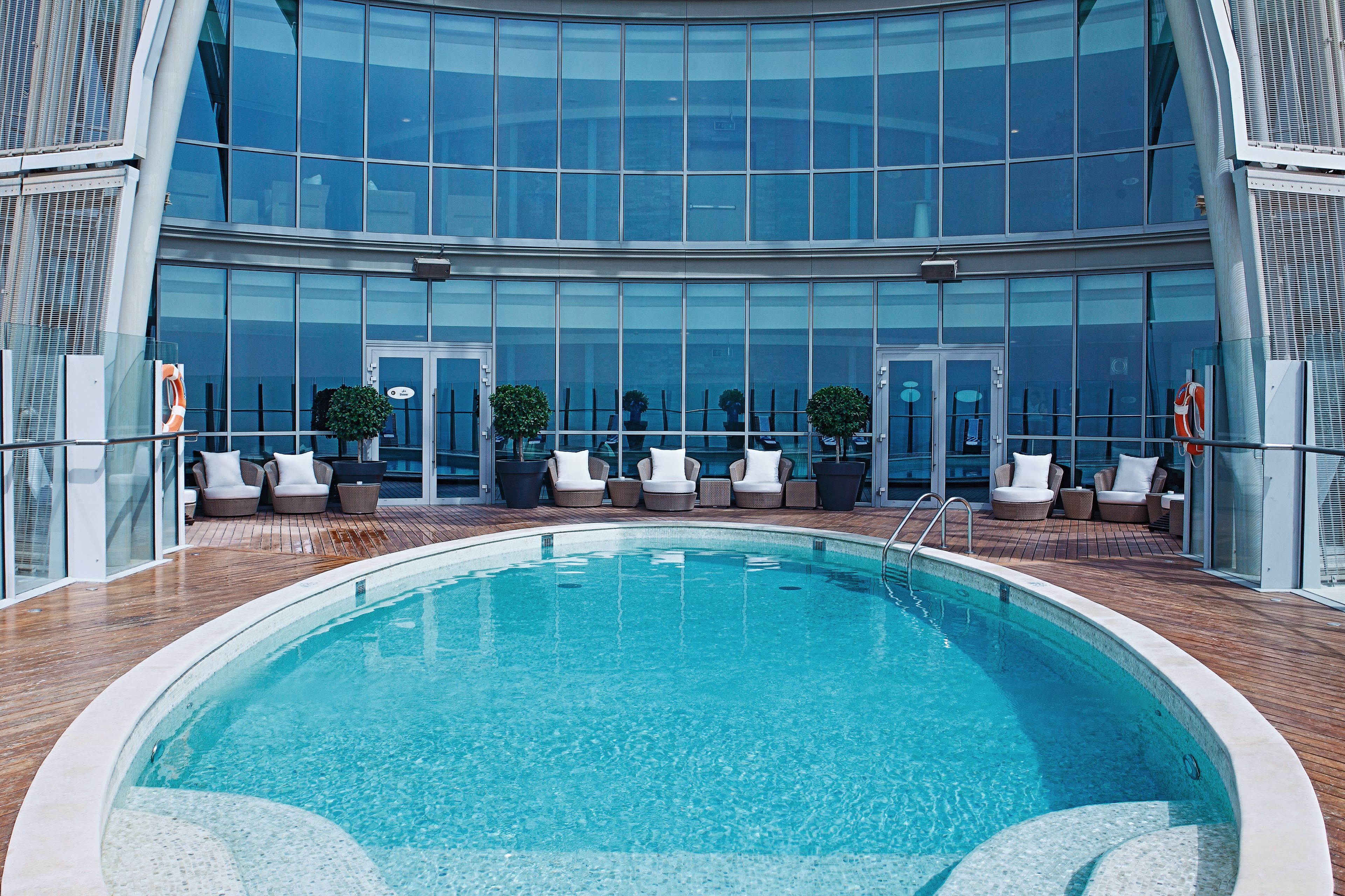 Área de piscina com espreguiçadeiras e plantas em vasos, cercada por paredes de vidro e uma estrutura de edifício moderna.