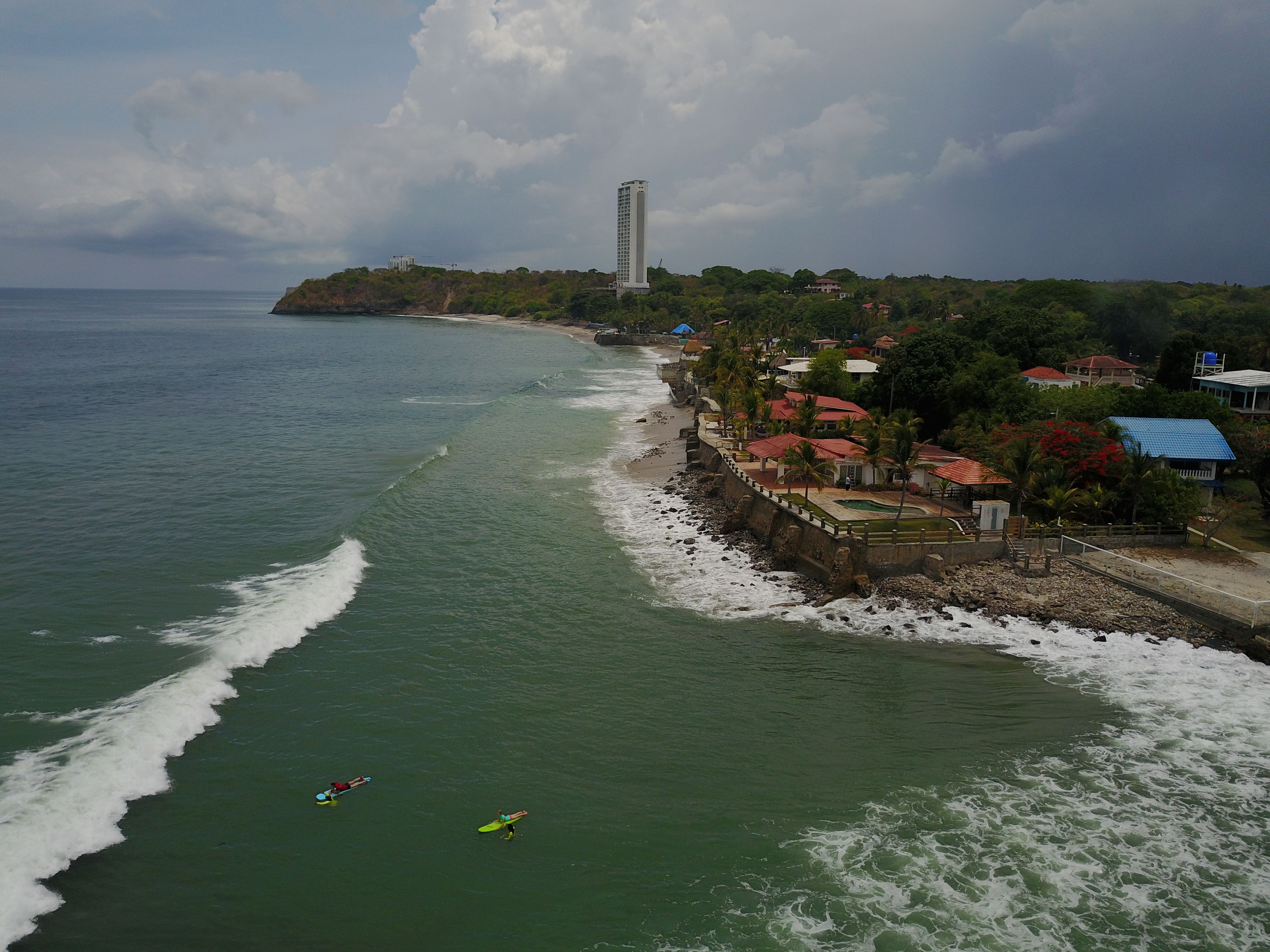 Uma área costeira com uma praia, um edifício alto e uma fileira de casas com telhados vermelhos.