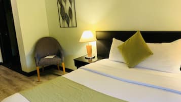 Quarto de hotel com uma cama, uma cadeira, mesa de cabeceira com abajur e uma obra de arte na parede.