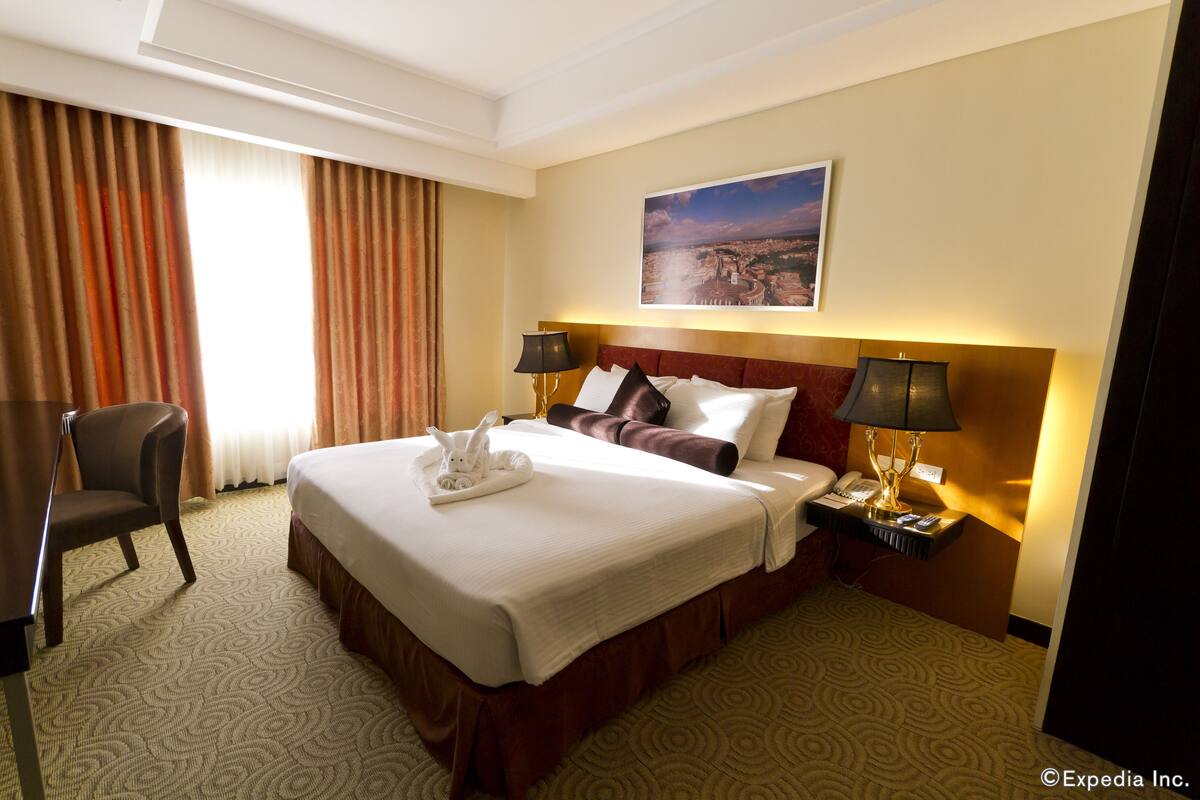 Quarto de hotel com uma cama grande, duas mesas de cabeceira com luminárias, uma cadeira e um quadro na parede.