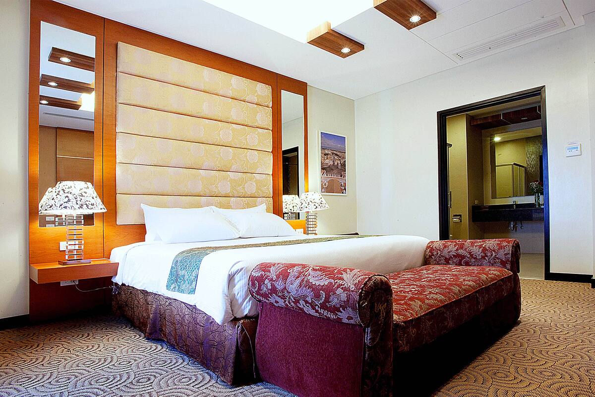Quarto de hotel com uma cama grande, cabeceira de madeira, duas luminárias de apoio, um banco estofado vermelho e um tapete estampado.
