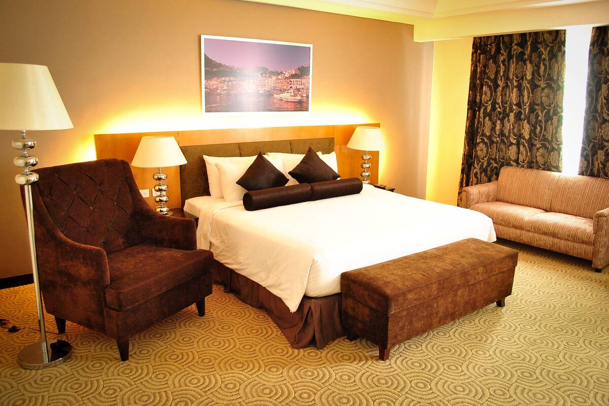 Quarto de hotel com uma cama grande, duas luminárias de cabeceira, uma poltrona marrom, um sofá e um tapete estampado.