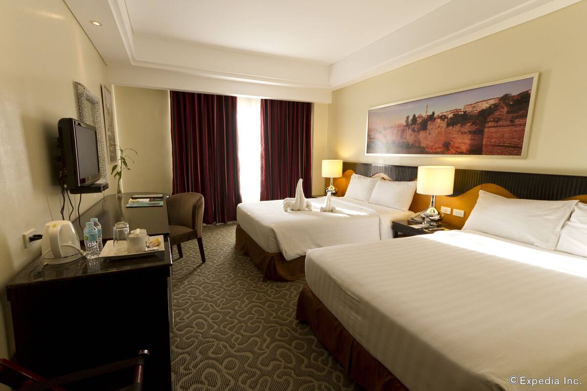 Quarto de hotel com duas camas, uma escrivaninha com computador, uma cadeira, televisão e um quadro na parede.