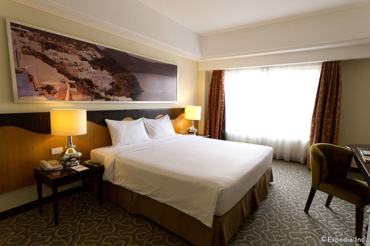 Quarto de hotel com uma cama grande, duas luminárias de cabeceira, uma mesa com cadeira, um quadro grande na parede e uma janela com cortinas.