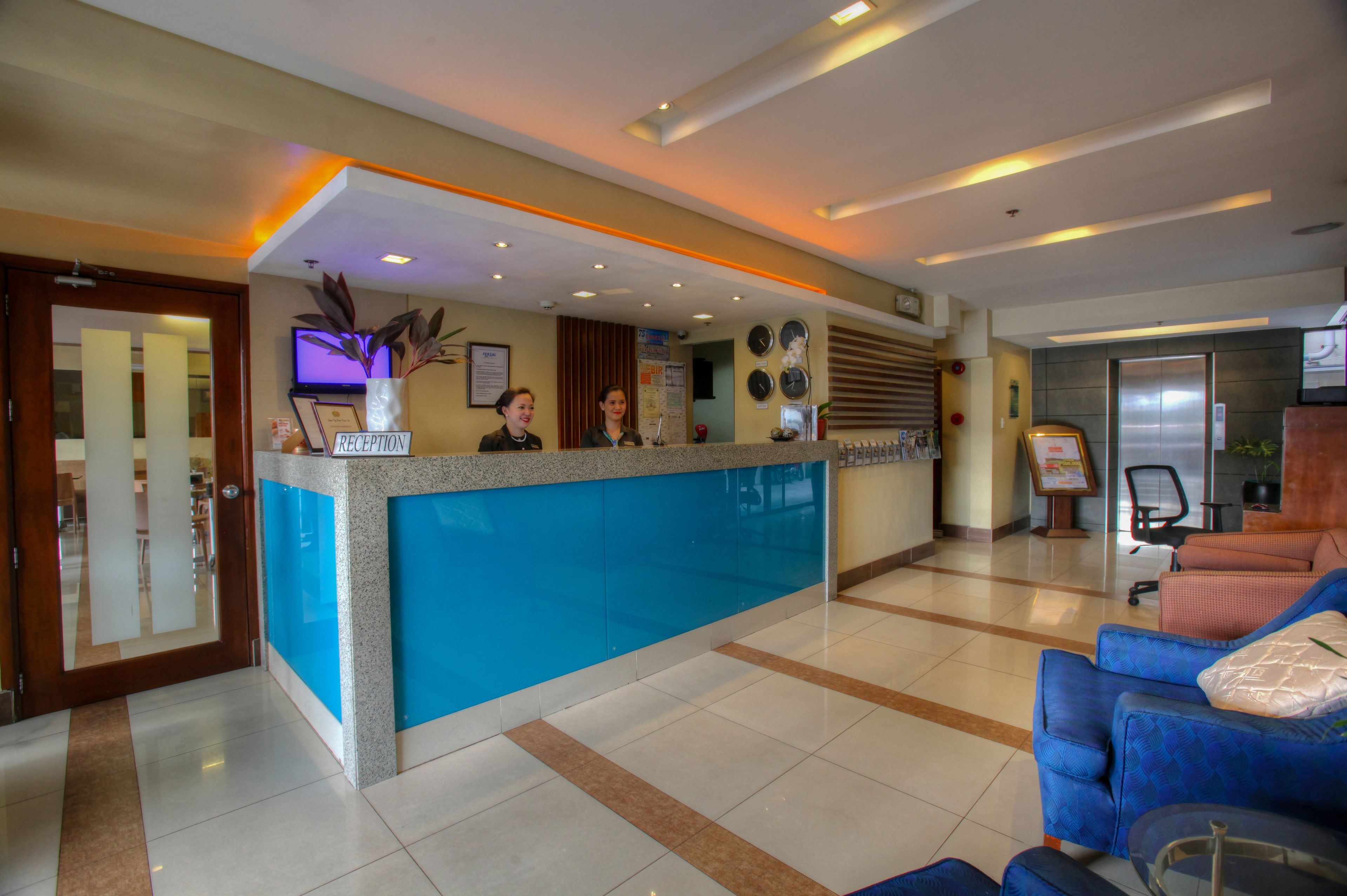 Uma área de recepção de hotel com um balcão azul, dois recepcionistas, um relógio e uma área de estar com sofás azuis.