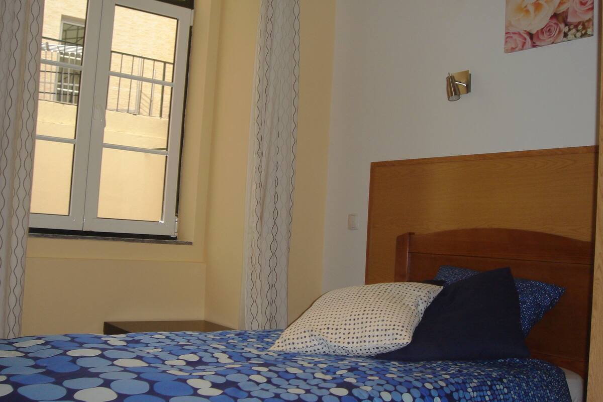 Quarto com cama, janela com cortinas e interruptor de luz na parede.