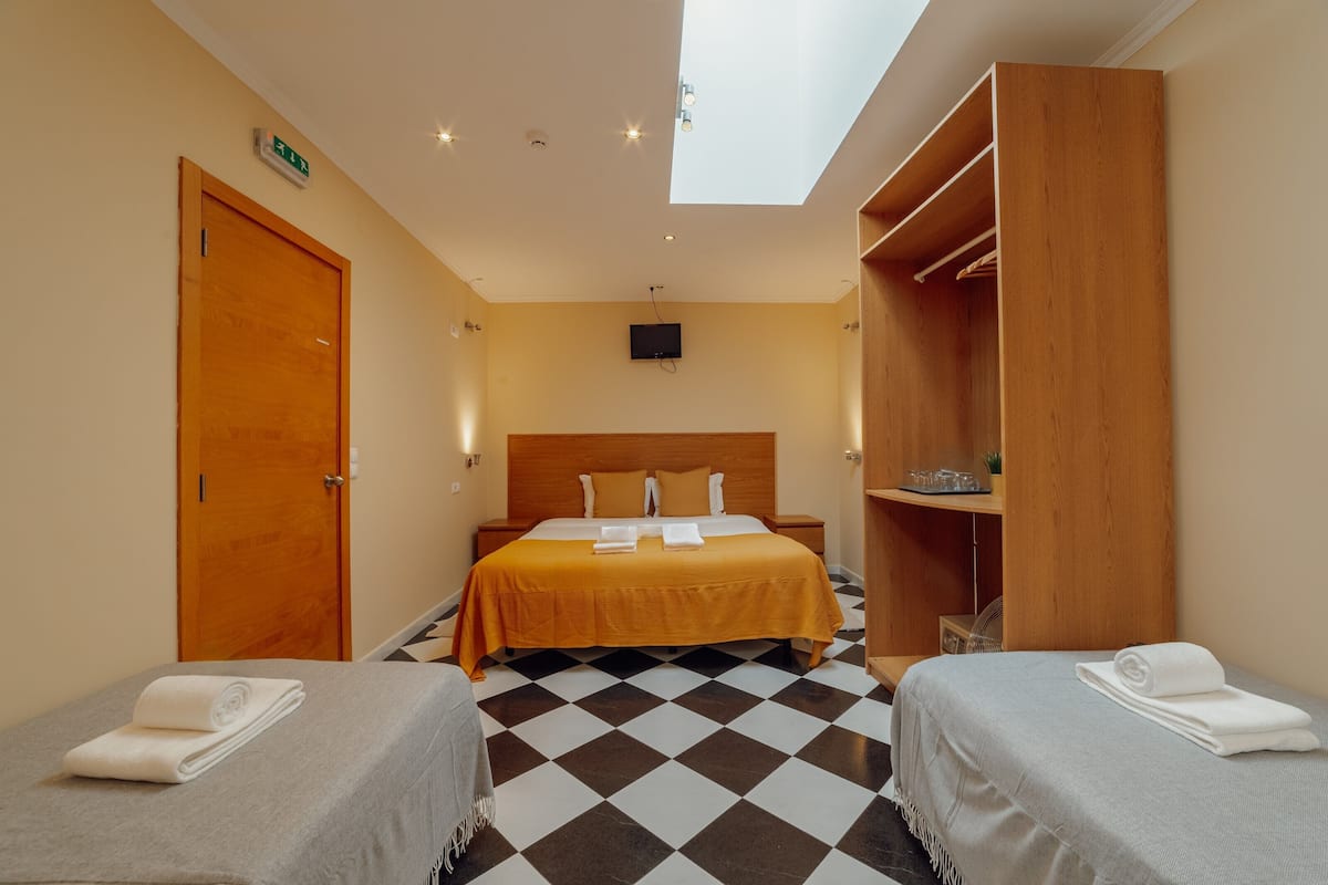 Quarto de hotel com uma cama, duas mesas de cabeceira, um guarda-roupa, piso xadrez e um claraboia.