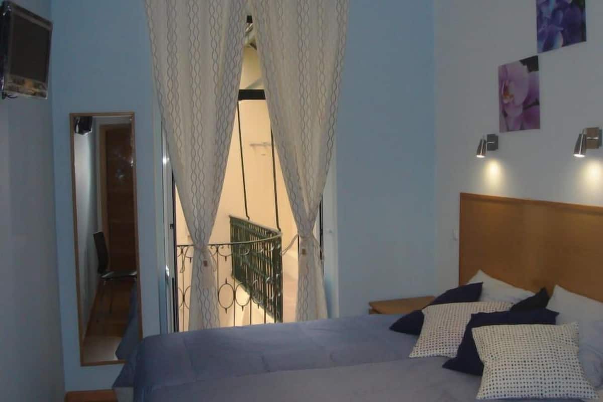 Quarto com cama, cortinas, cabeceira, espelho e vista para uma varanda.