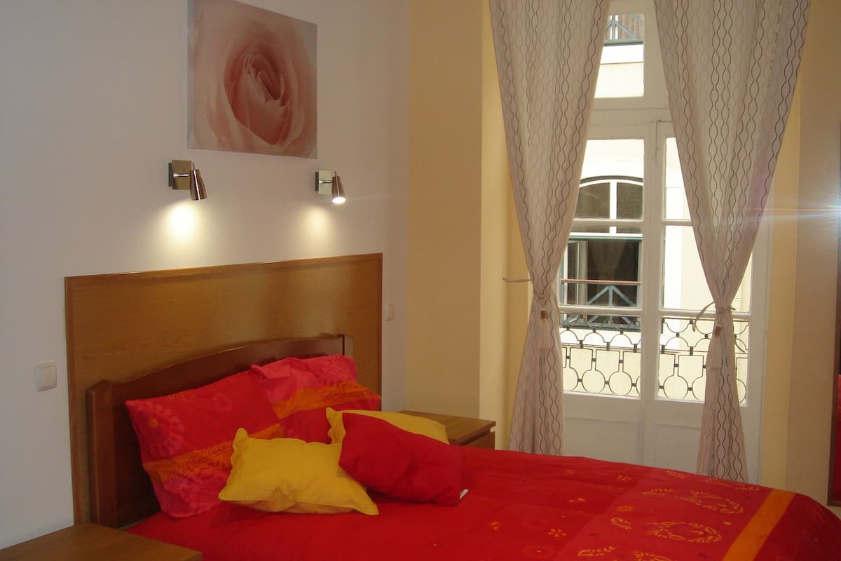 Cama bem arrumada com colcha vermelha e amarela, cabeceira de madeira e duas luminárias de mesa.