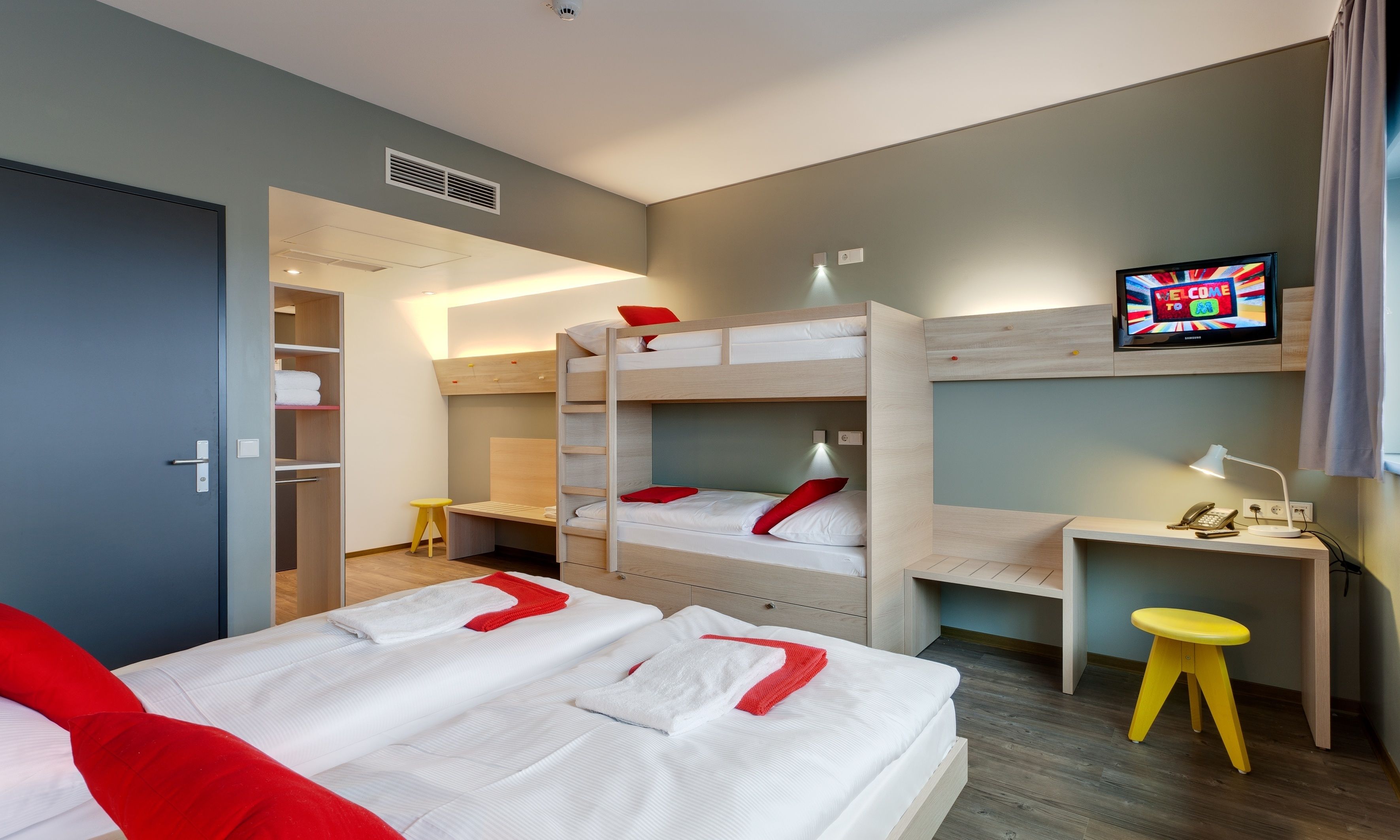 Quarto de hotel moderno com beliches, escrivaninha, cadeira, televisão e abajur.
