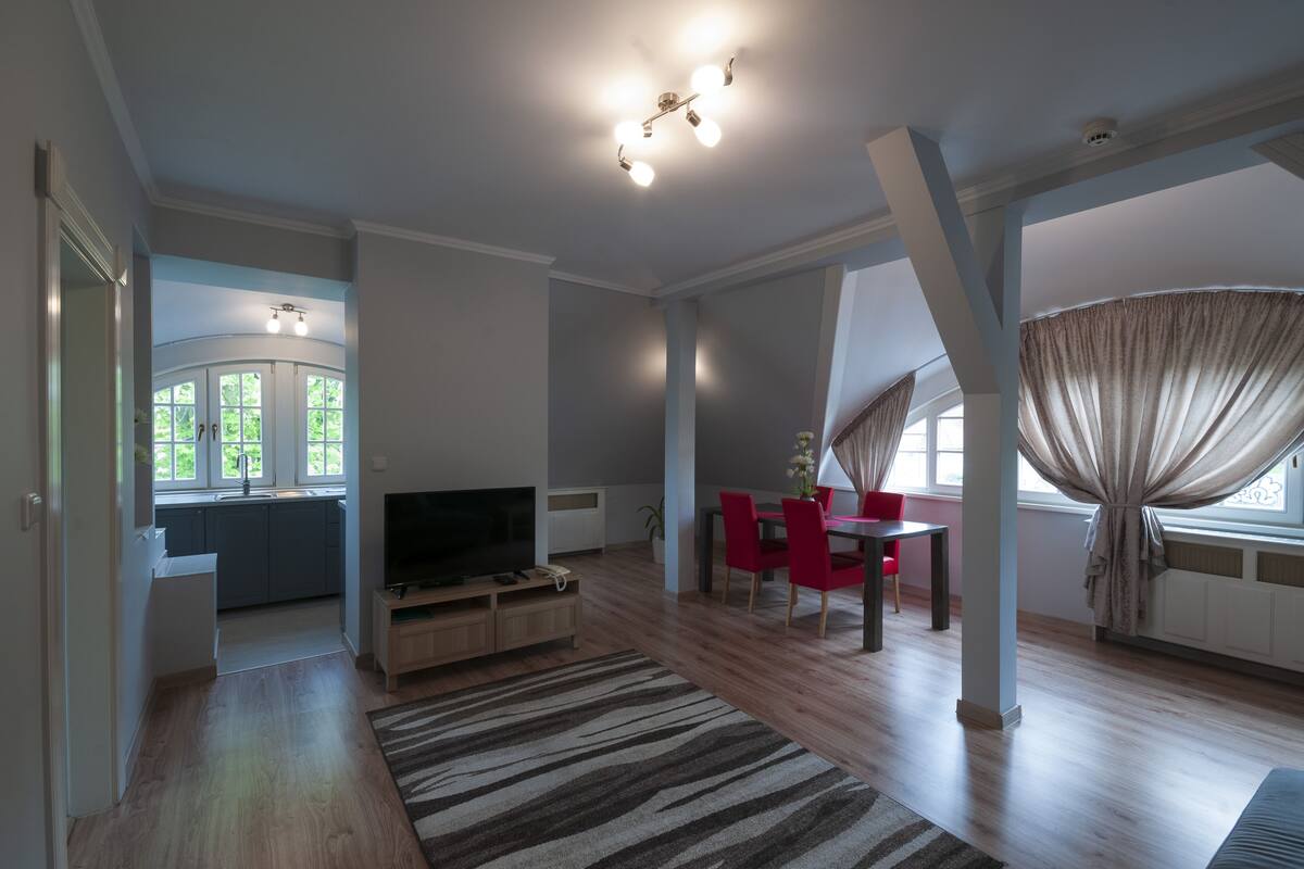 Uma sala de estar moderna com televisão, mesa de jantar e cadeiras vermelhas. Há uma área de cozinha com janela e pia.