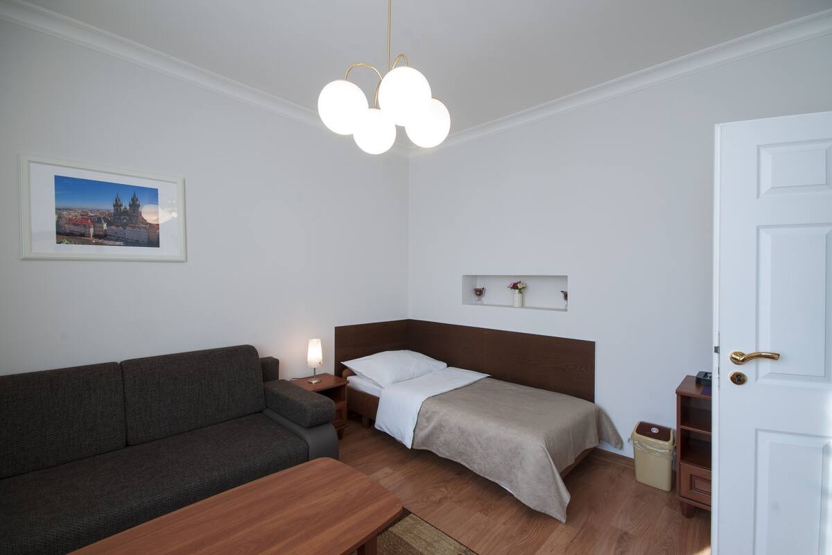 Quarto pequeno e bem arrumado com uma cama, um sofá, uma mesinha de cabeceira com abajur, um quadro na parede e uma porta.