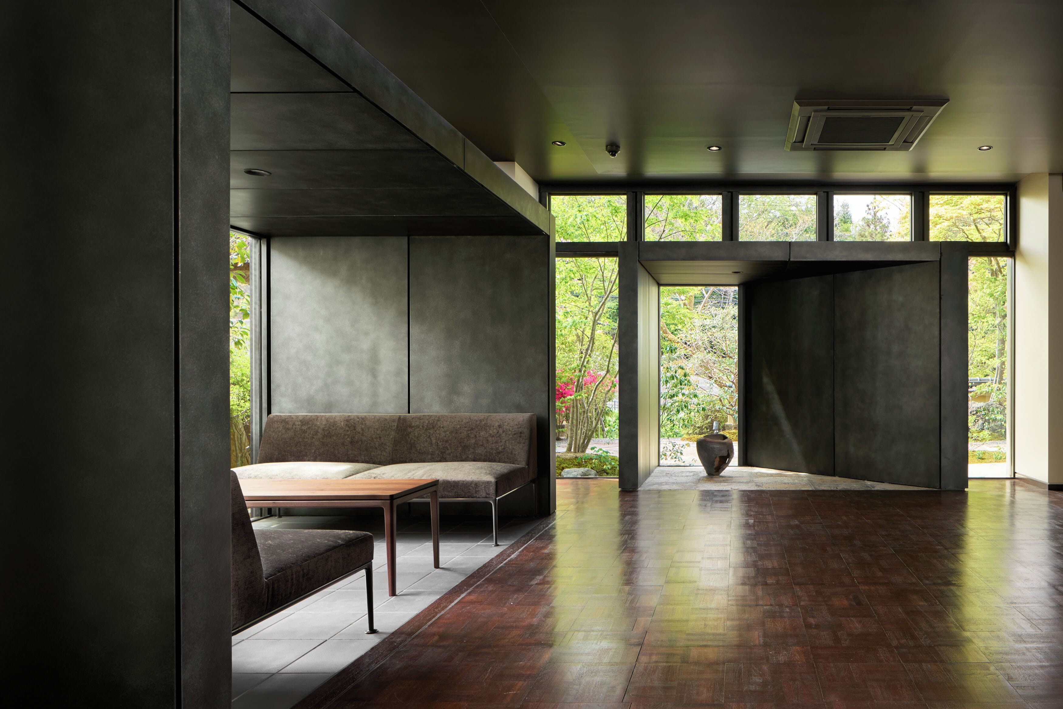 Um interior moderno com um sofá em tons escuros, uma mesa de madeira e uma janela grande com vista para o verde.