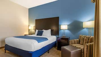 Quarto de hotel com uma cama grande, duas mesas de cabeceira com abajures, uma poltrona estampada e um banquinho marrom.