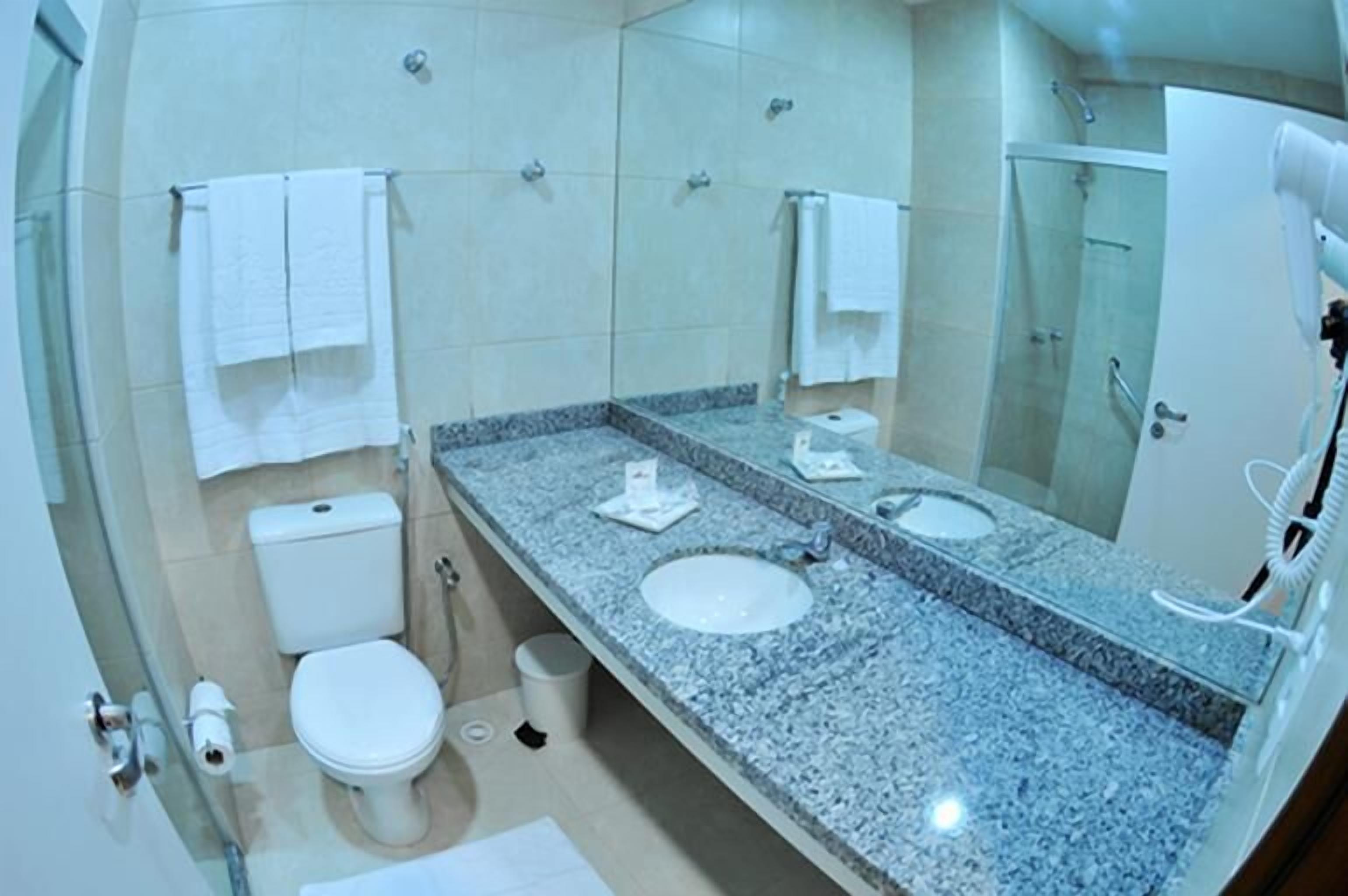Um banheiro moderno com bancada de granito, duas pias, vaso sanitário e box de vidro.