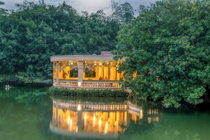 Uma casa flutuante isolada, iluminada e rodeada por vegetação densa, refletindo em águas calmas.