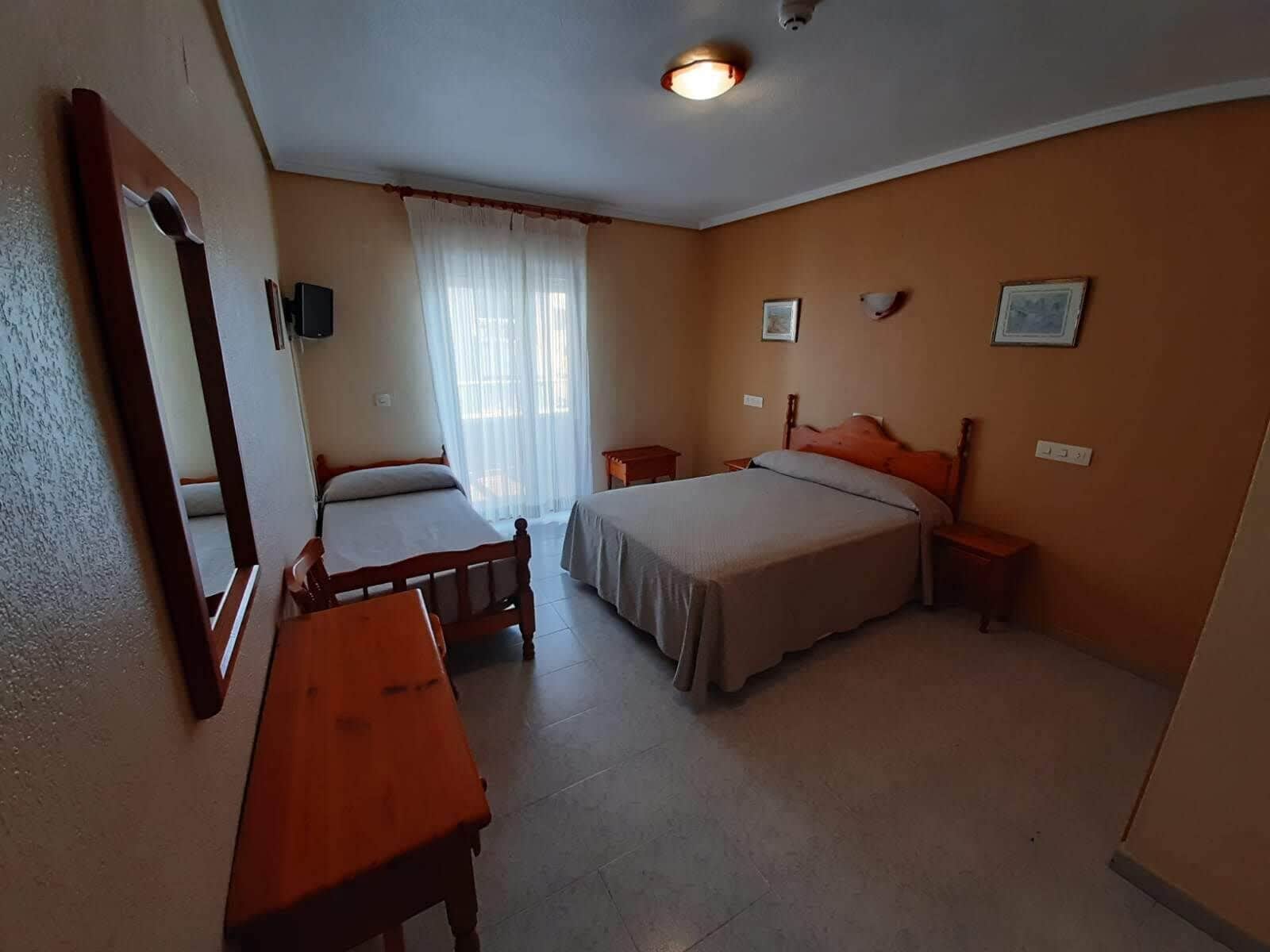 Quarto de hotel com duas camas de solteiro, uma escrivaninha de madeira, um espelho e uma luminária no teto.
