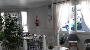 Quarto de hotel moderno com área de jantar, uma grande planta em vaso, um relógio e vista para a rua.