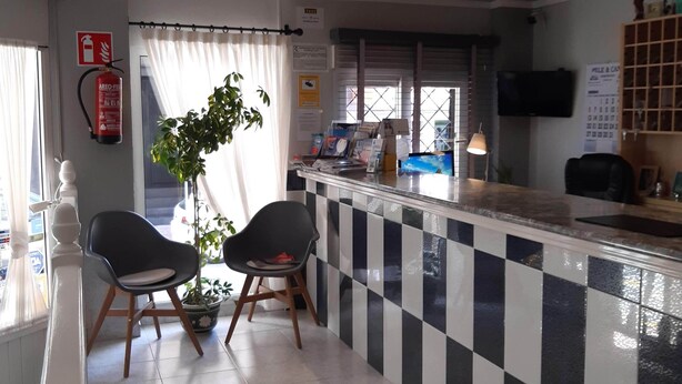 Um saguão de hotel com piso quadriculado em preto e branco, duas cadeiras pretas, um balcão de recepção, um extintor de incêndio e um calendário na parede.