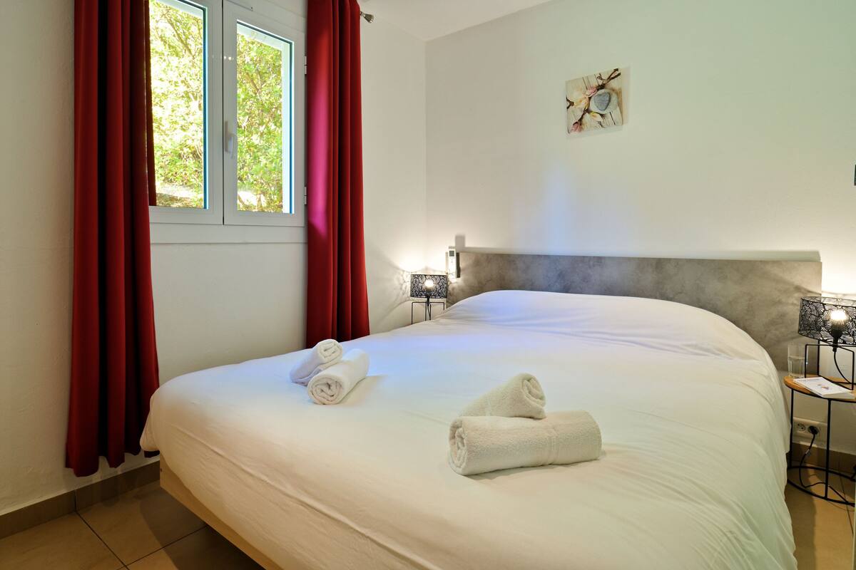 Cama bem arrumada com lençóis e toalhas brancas, cabeceira, mesa de cabeceira com abajur e janela com cortinas vermelhas.